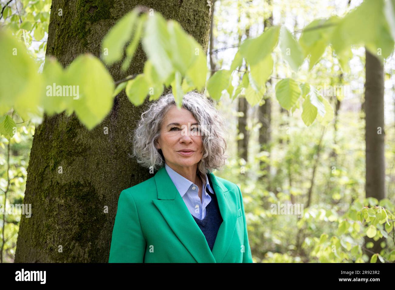 Senior woman leaning on tree in forest wearung green coat Stock Photo ...