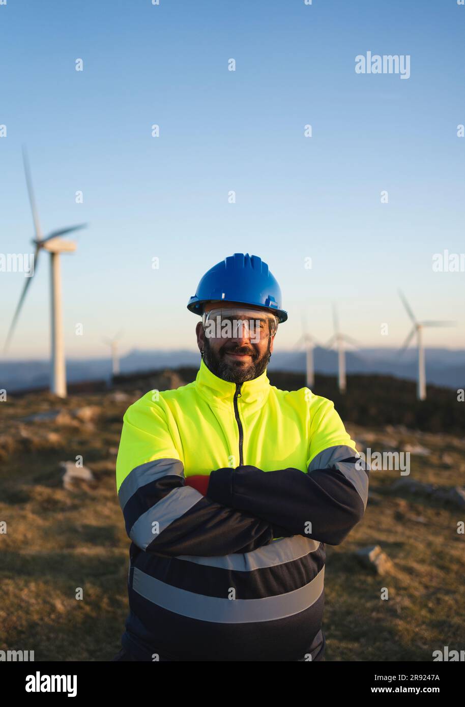 Smiling engineer with arms crossed standing in front of wind turbines ...
