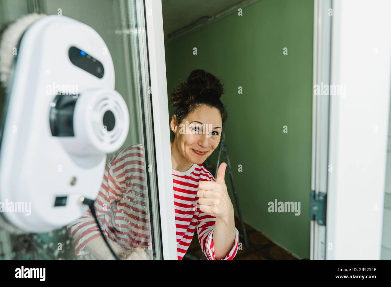 Happy woman showing thumbs up gesture and cleaning window with robot ...