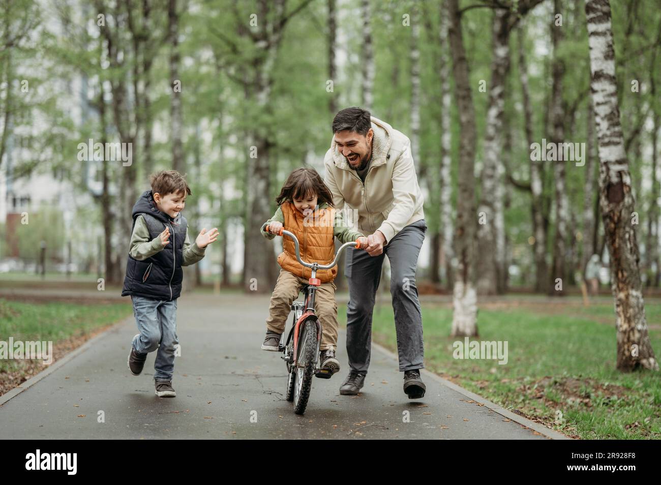Boy cheering brother learning to ride bicycle with father at park Stock ...