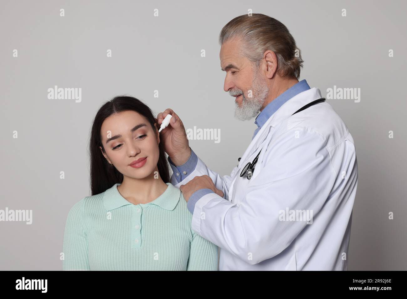 Doctor dripping medication into woman's ear on light grey background ...