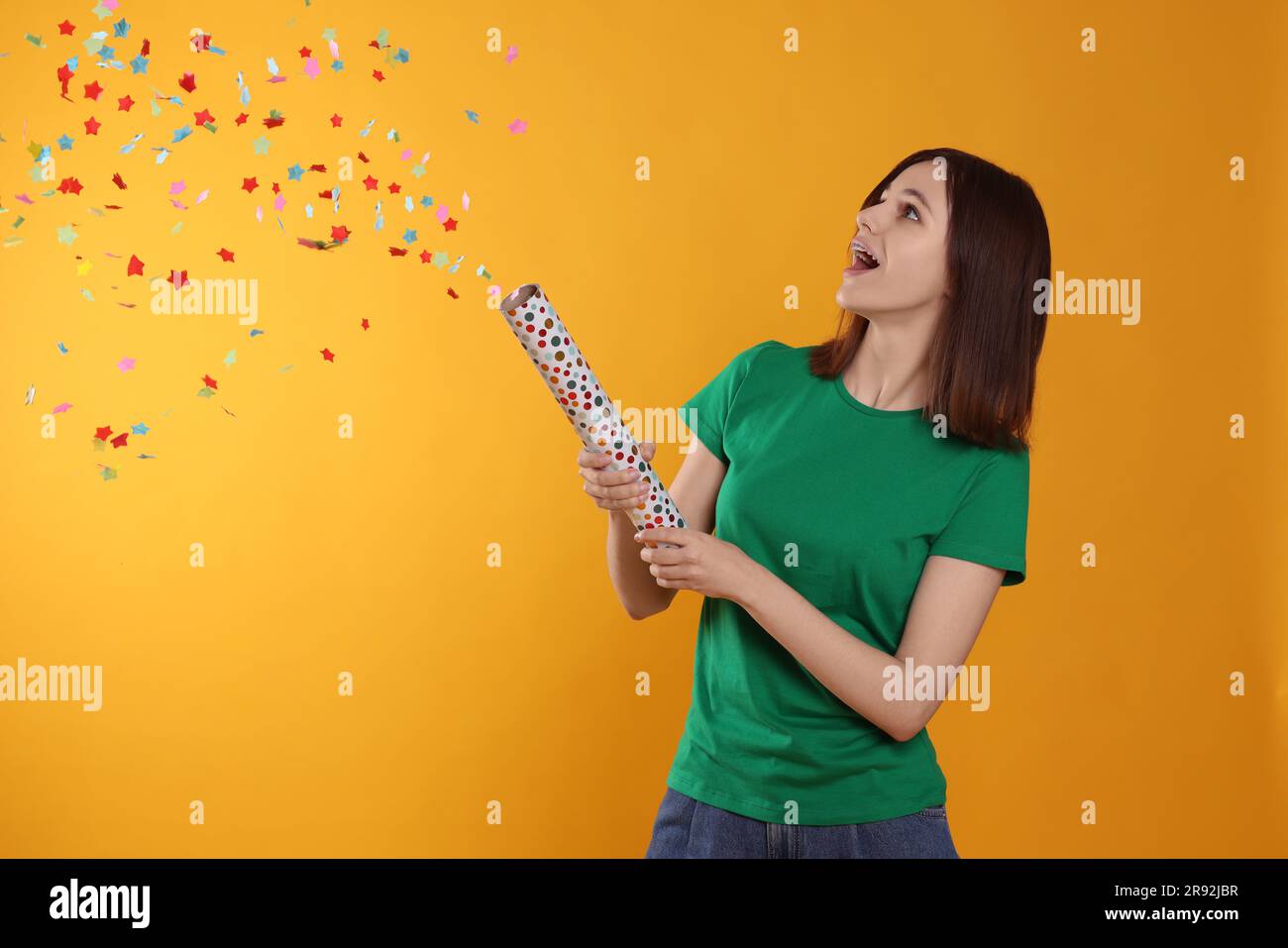 Happy teenage girl blowing up party popper on orange background Stock ...