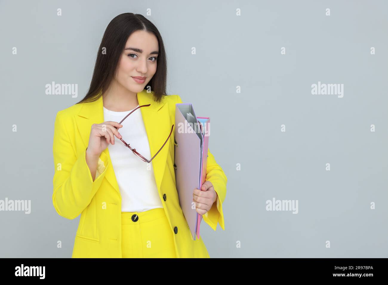 Young female intern with eyeglasses and folders on grey background ...