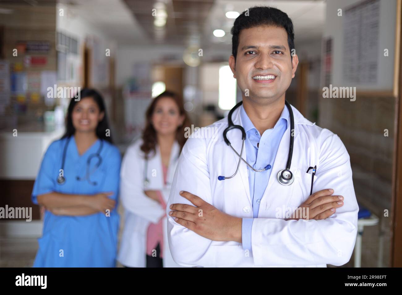 Confident doctor with colleague standing in background at hospital corridor. Wearing lab coat ...