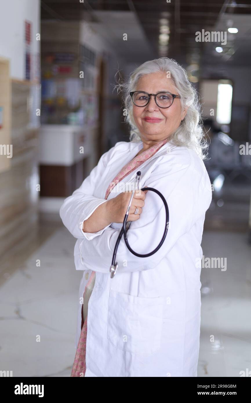 Portrait of senior female doctor with arms crossed standing in hospital corridor. Happy Indian ...