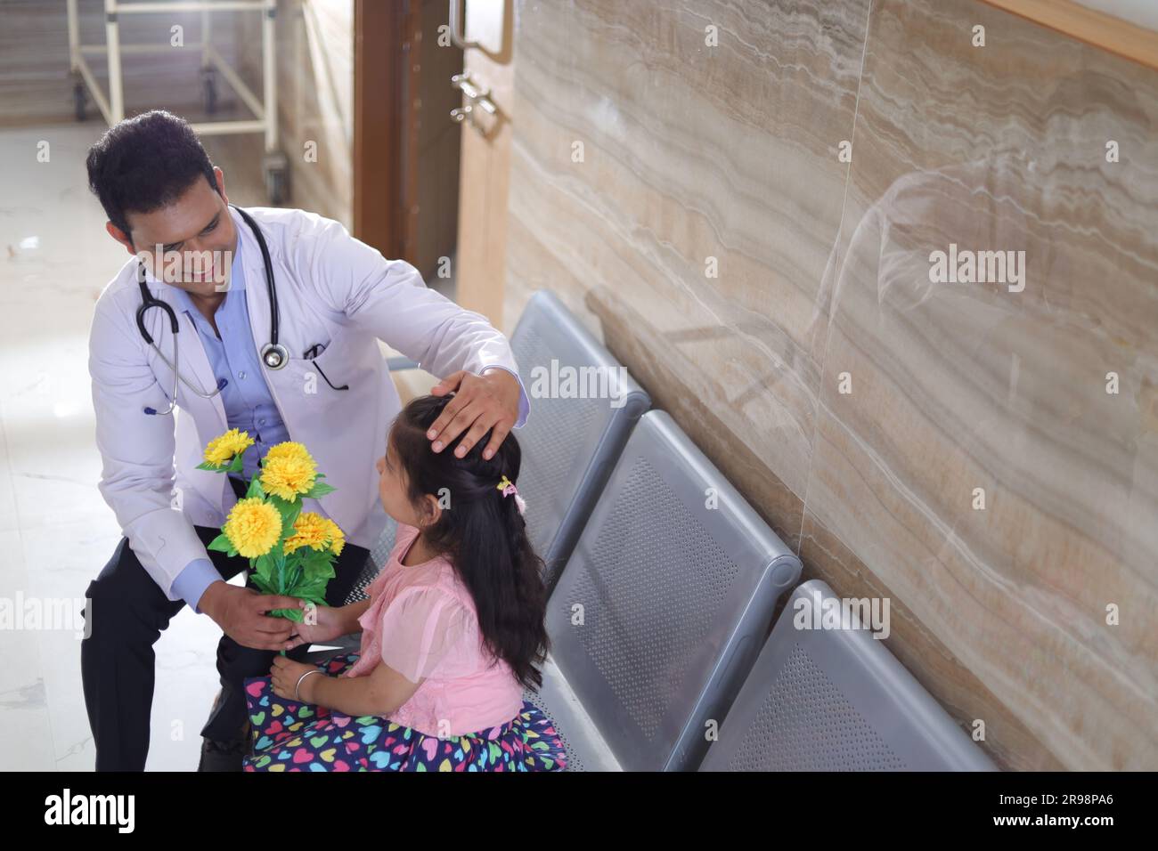 Happy girl child sitting on bench in hospital corridor, senior doctor giving her yellow flowers ...