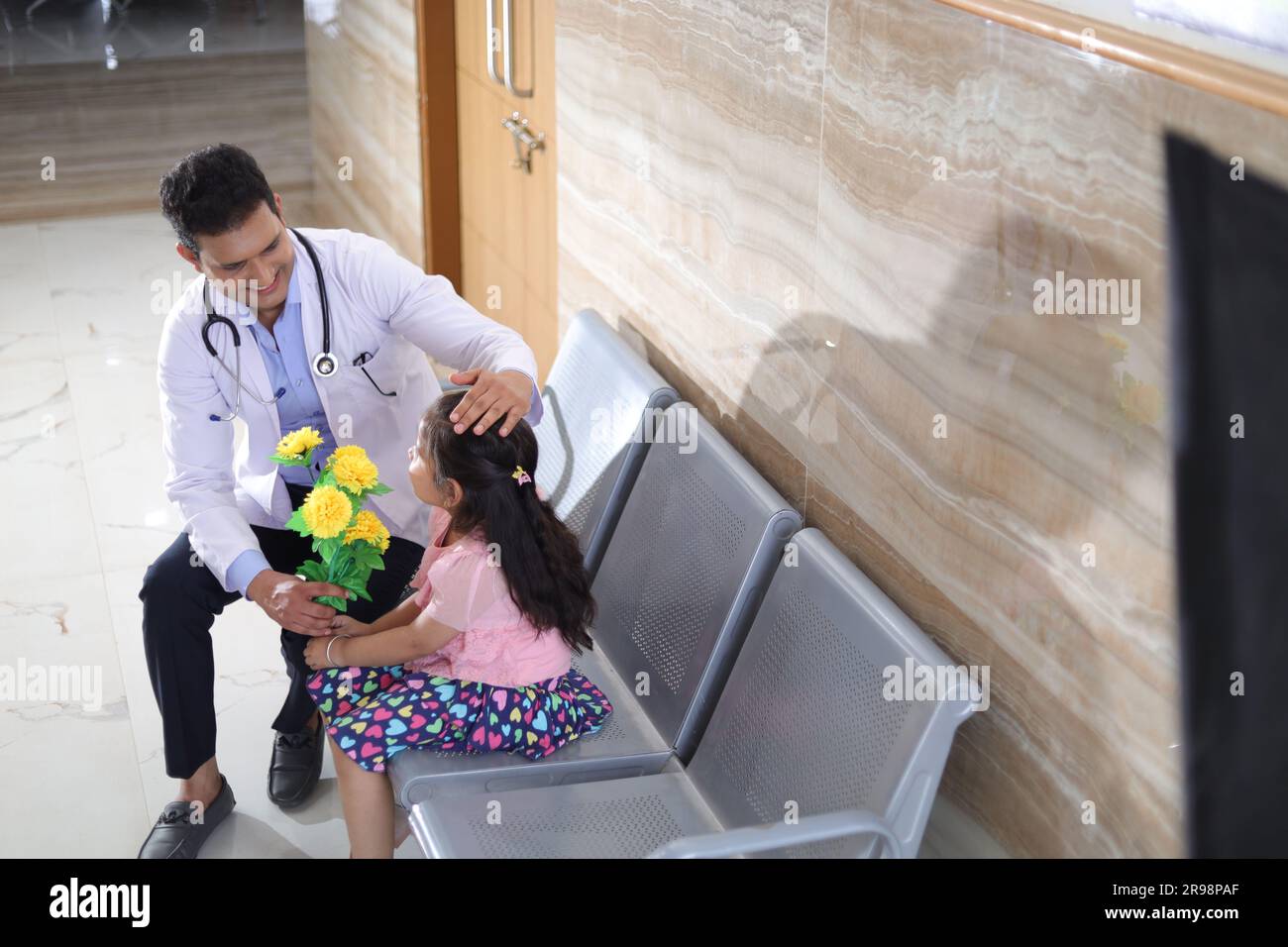 Happy girl child sitting on bench in hospital corridor, senior doctor giving her yellow flowers ...