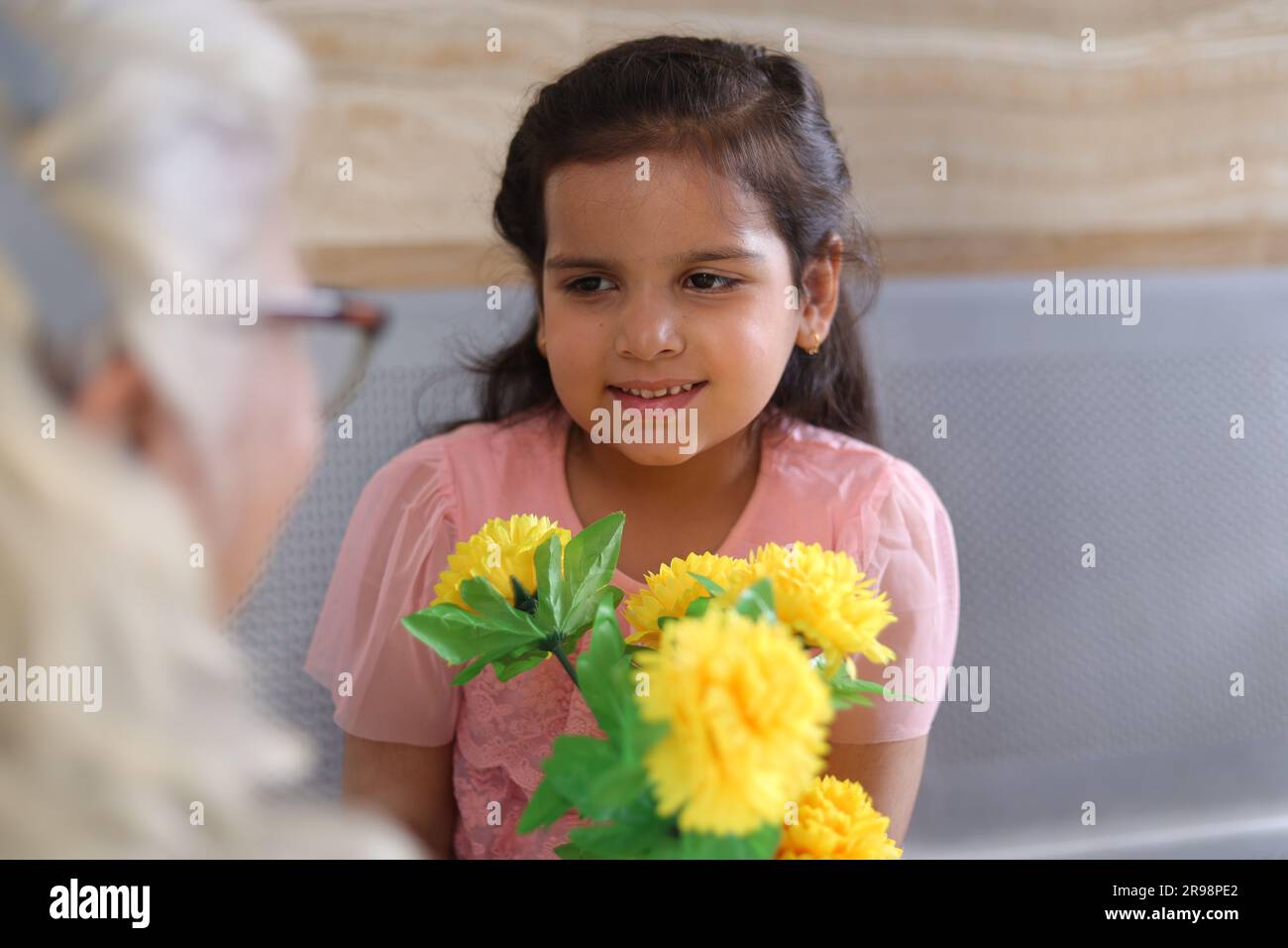 Happy girl child sitting on bench in hospital corridor, senior doctor giving her yellow flowers ...
