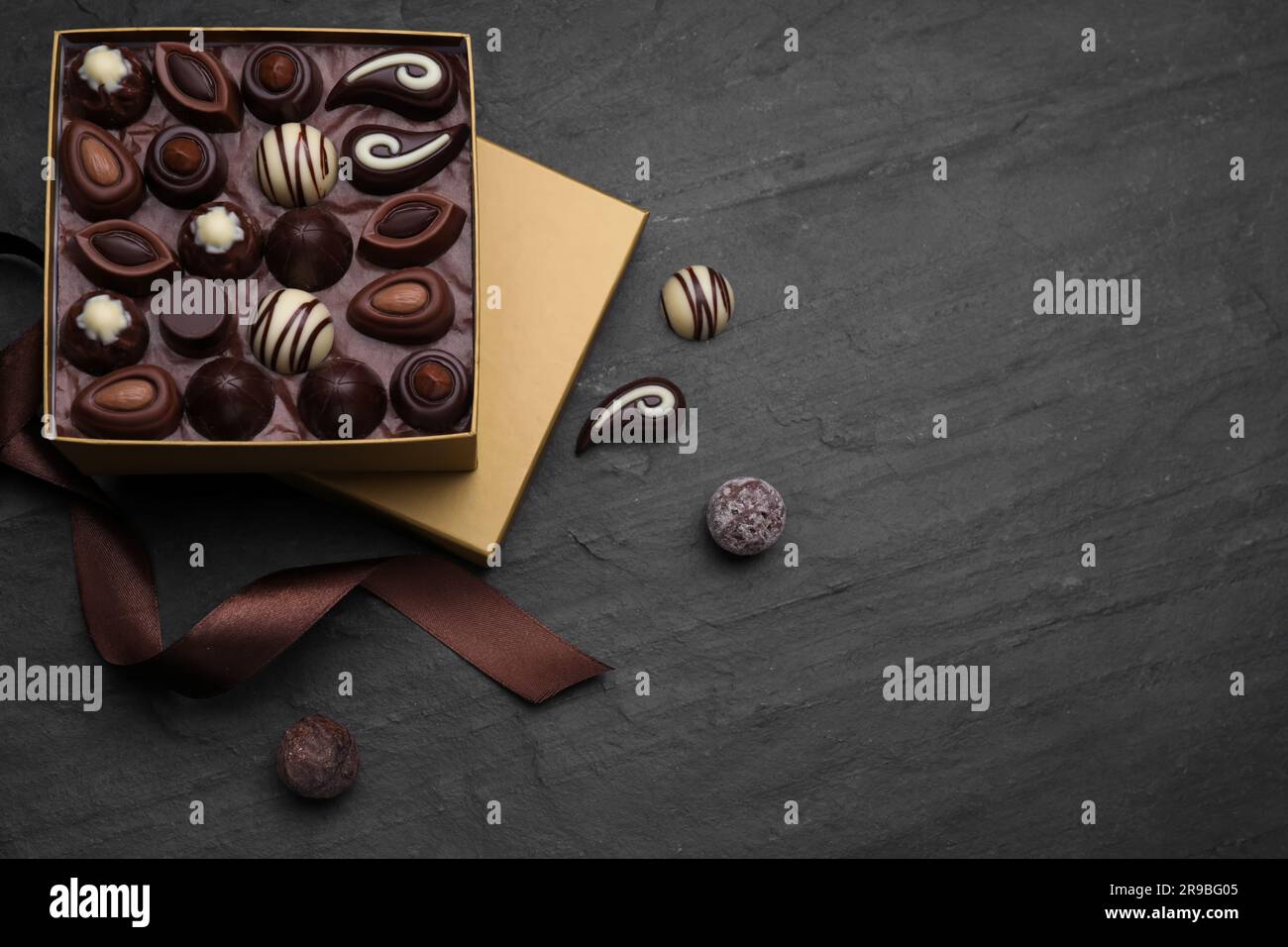 Open box of delicious chocolate candies and brown ribbon on black table, flat lay. Space for ...