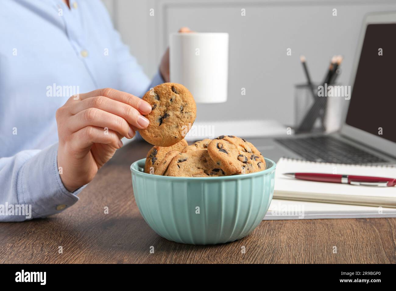Office worker with cup of drink taking chocolate chip cookie from bowl ...