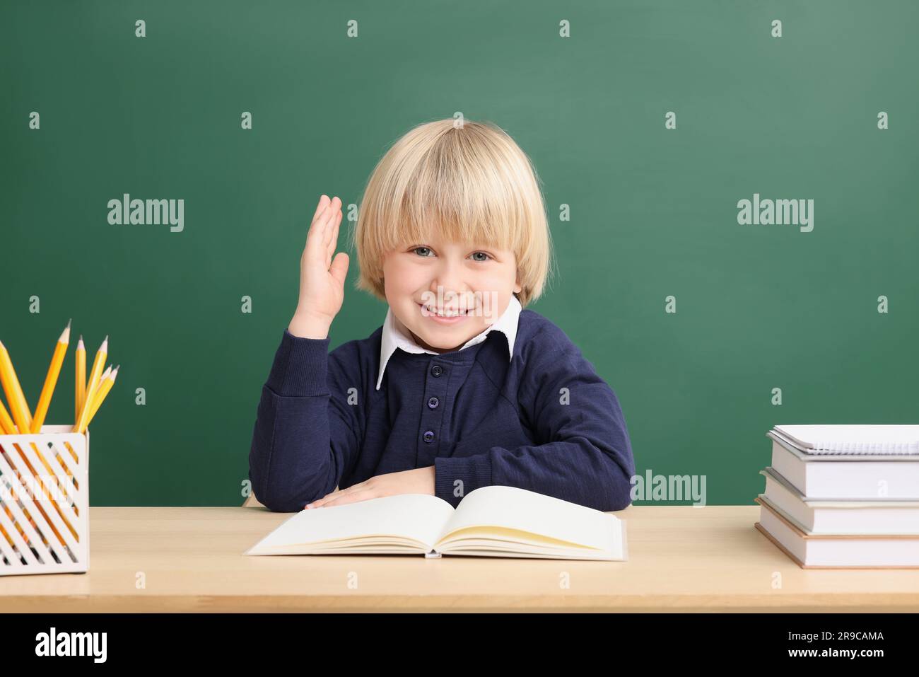 Happy little school child raising hand while sitting at desk with books ...