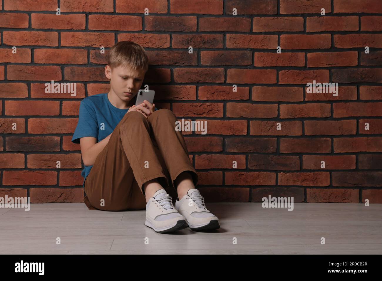 Boy with smartphone sitting on floor near brick wall, space for text ...