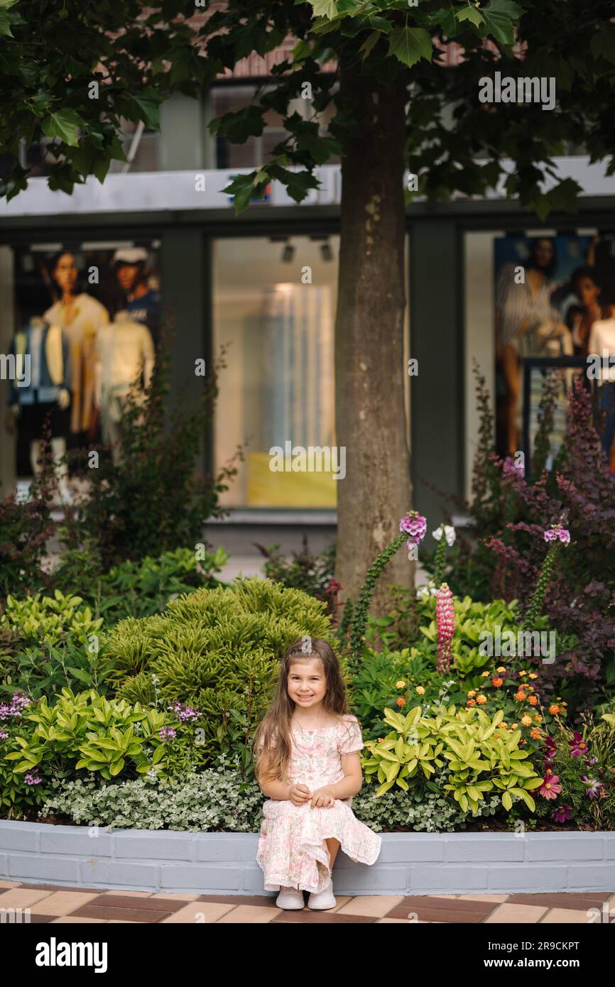 Cute little girl in gorgeous dress sits in front of flower bed with ...