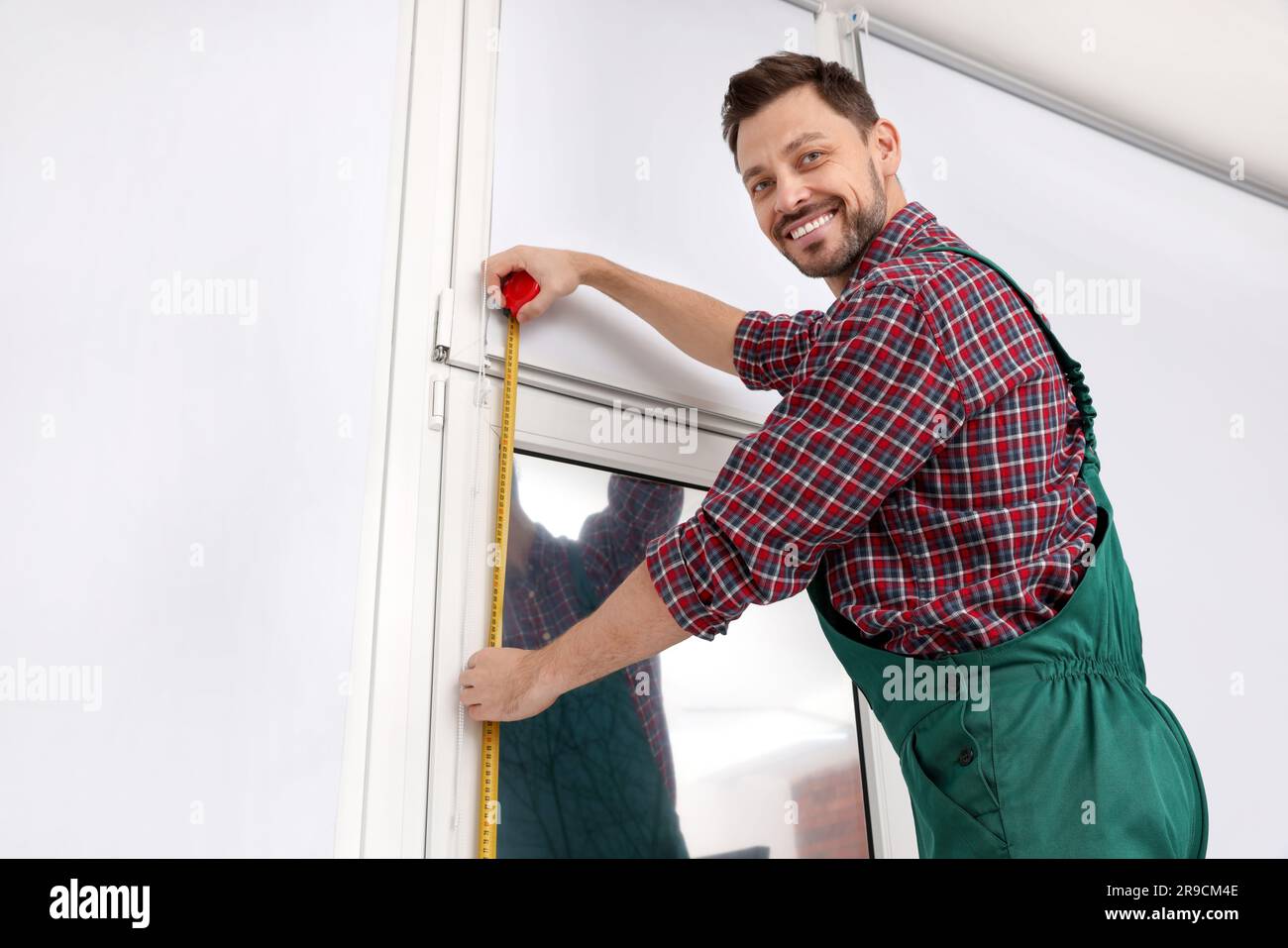 Worker in uniform using tape measure while installing roller window ...