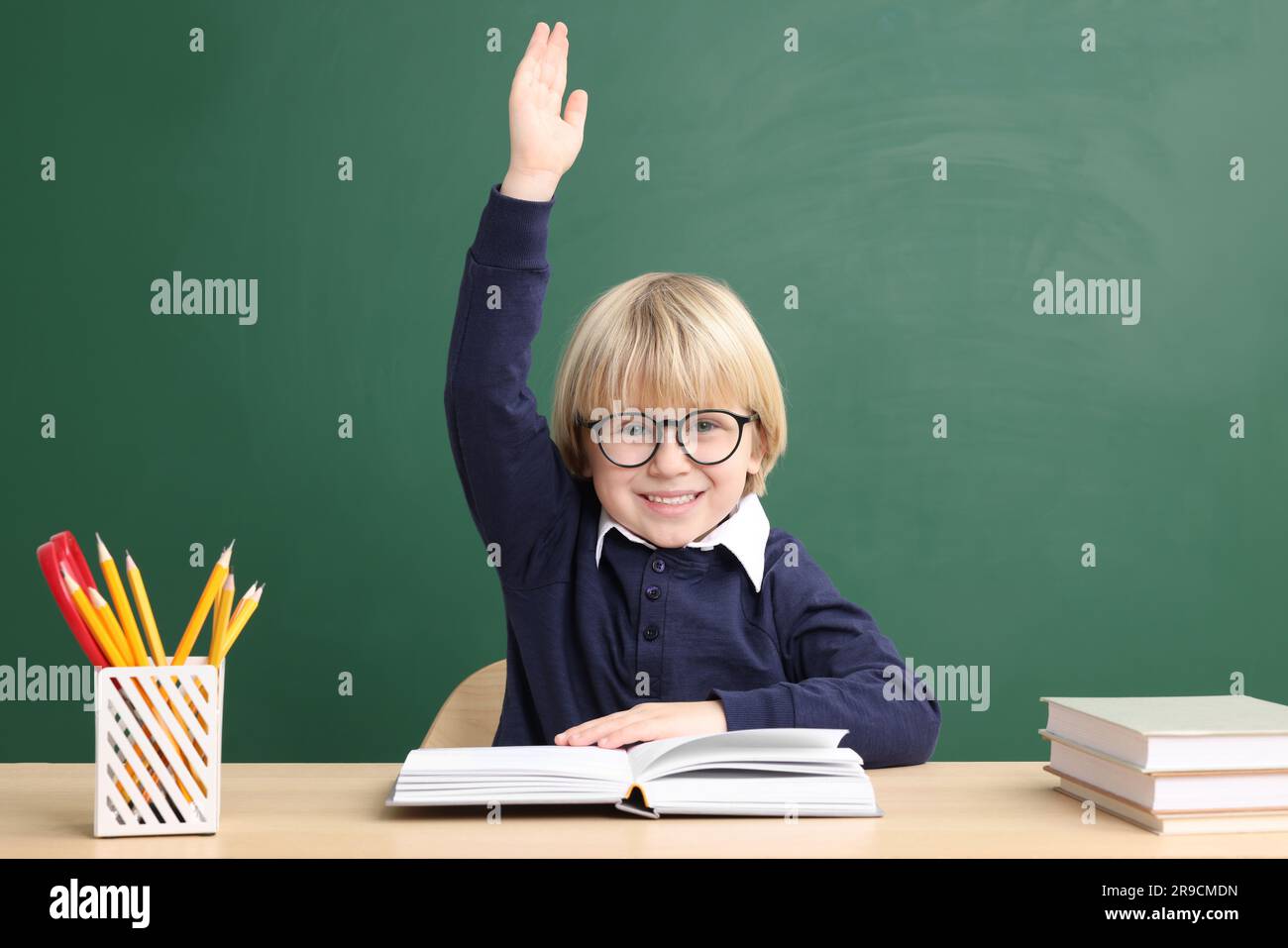 Happy little school child raising hand while sitting at desk with books ...