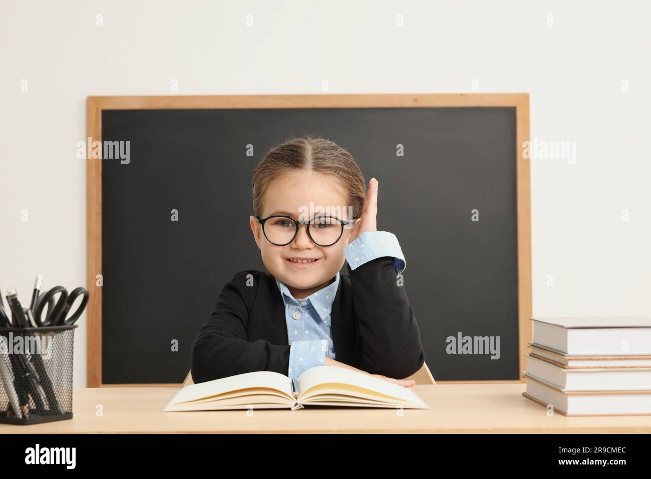 Happy little school child raising hand while sitting at desk with books ...