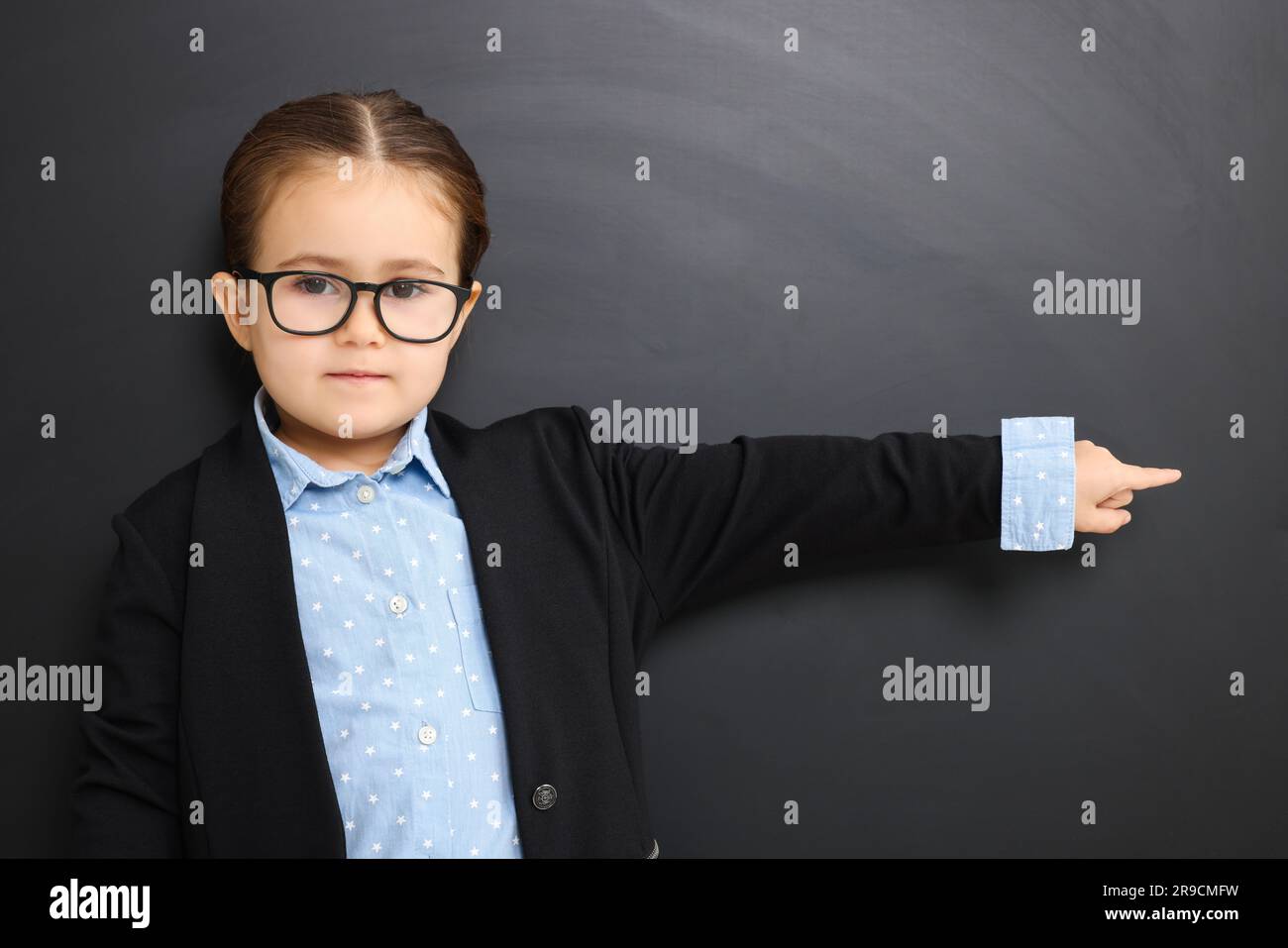 Little school child in uniform pointing at something near chalkboard ...