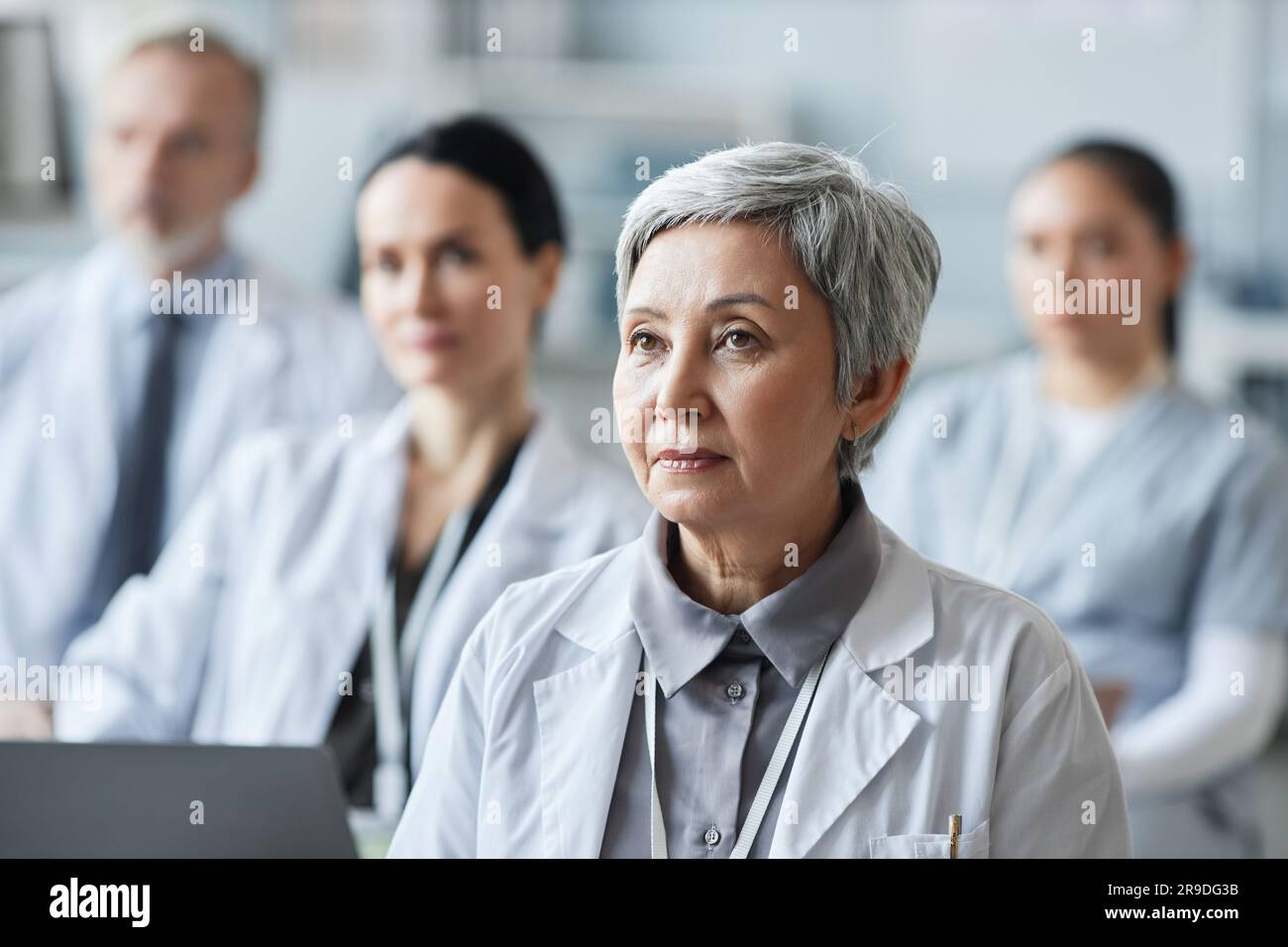 Mature Asian female doctor in lab coat looking at speaker making report ...