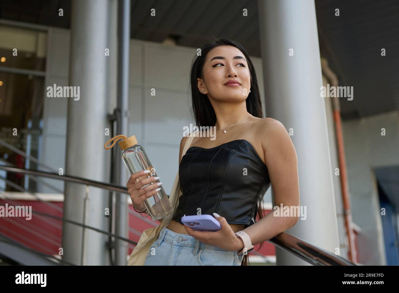 Beautiful Asian female posing outdoor with a glass bottle of water and modern smart phone in ...