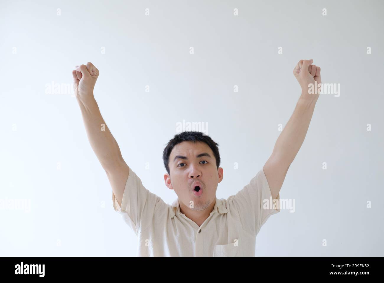 A young Asian man wearing a beige shirt is raising both hands with an ...