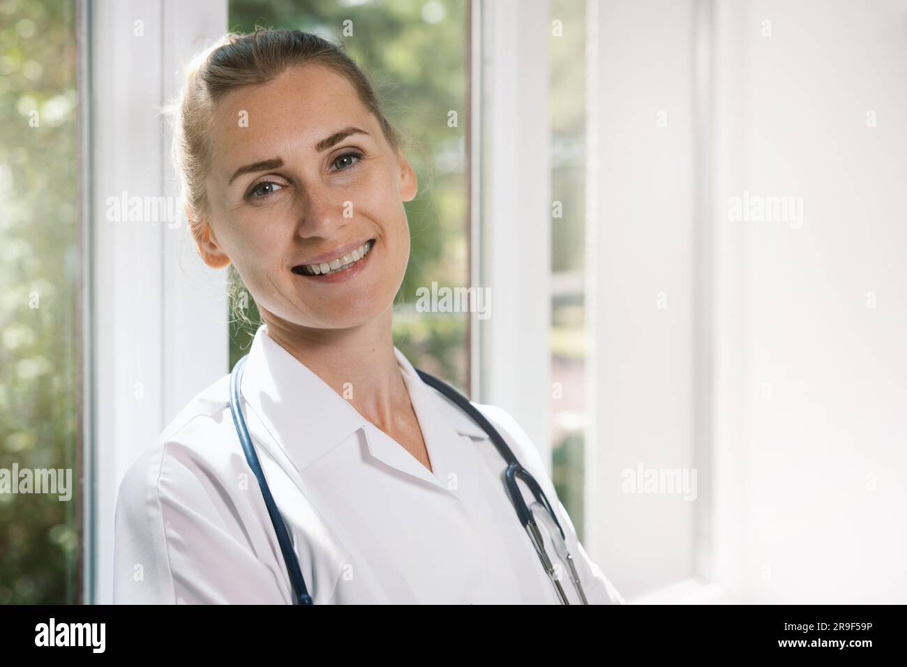 portrait of smiling female doctor with stethoscope standing by the window in hospital hallway ...