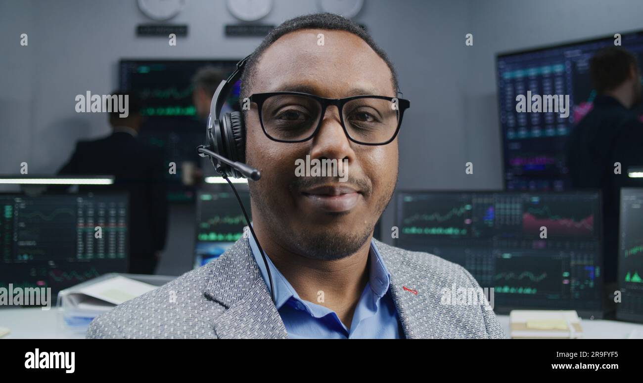 African American trader in headset sits at workplace, smiles and looks ...