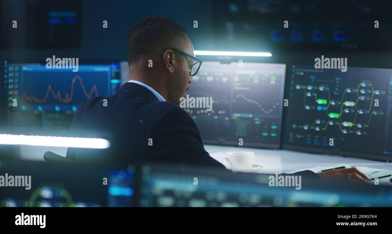 African American software engineer sits in front of computer and big digital screens with data ...
