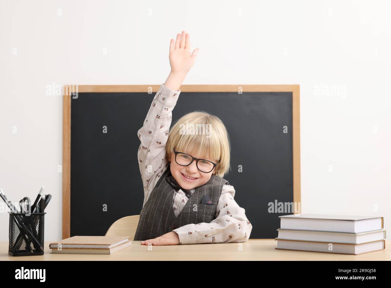 Happy little school child raising hand while sitting at desk with books ...