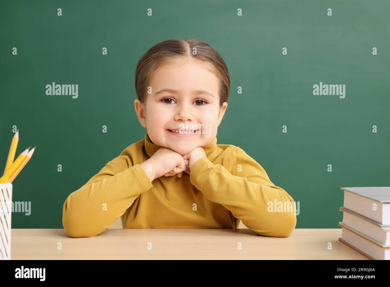 Happy little school child sitting at desk with books near chalkboard ...