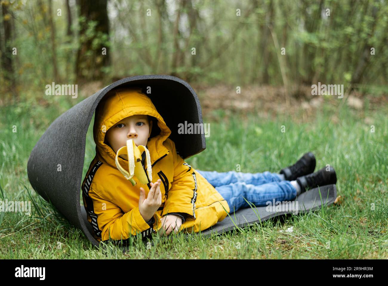 A young boy rests lying on a tourist mat in the woods and eats a banana ...