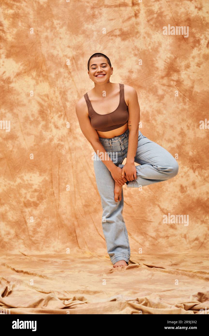 body positivity and confidence, happy young woman in crop top and jeans posing on mottled beige ...