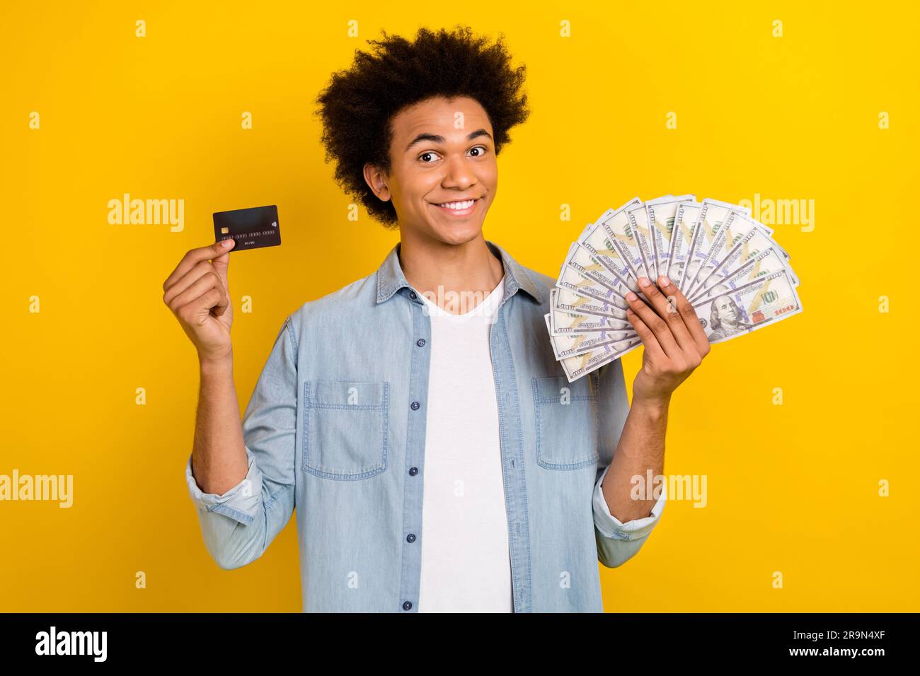 Photo of cute positive guy dressed denim shirt comparing bank card cash ...