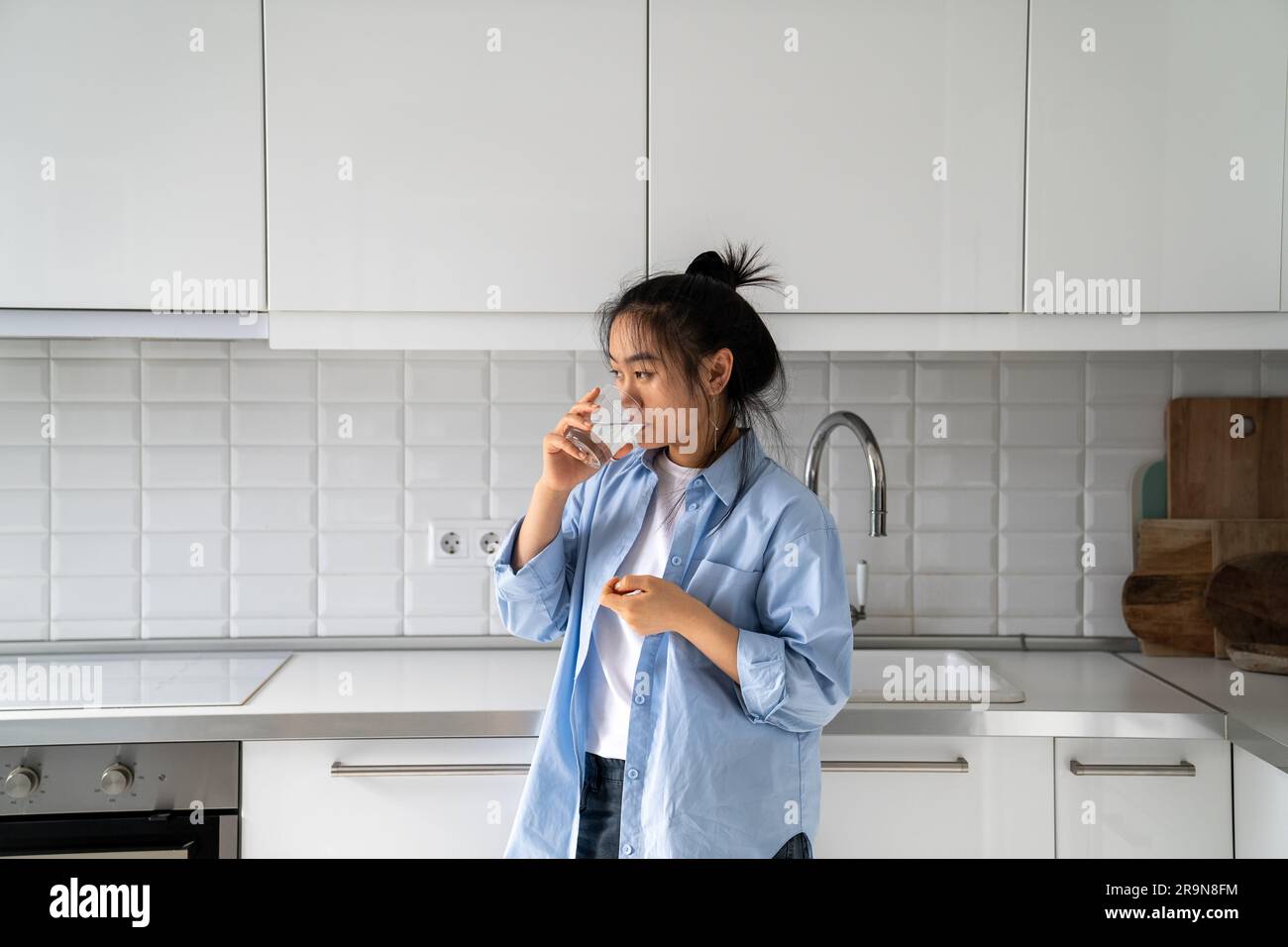 Weakened lonely Asian woman standing in kitchen of own home with glass ...