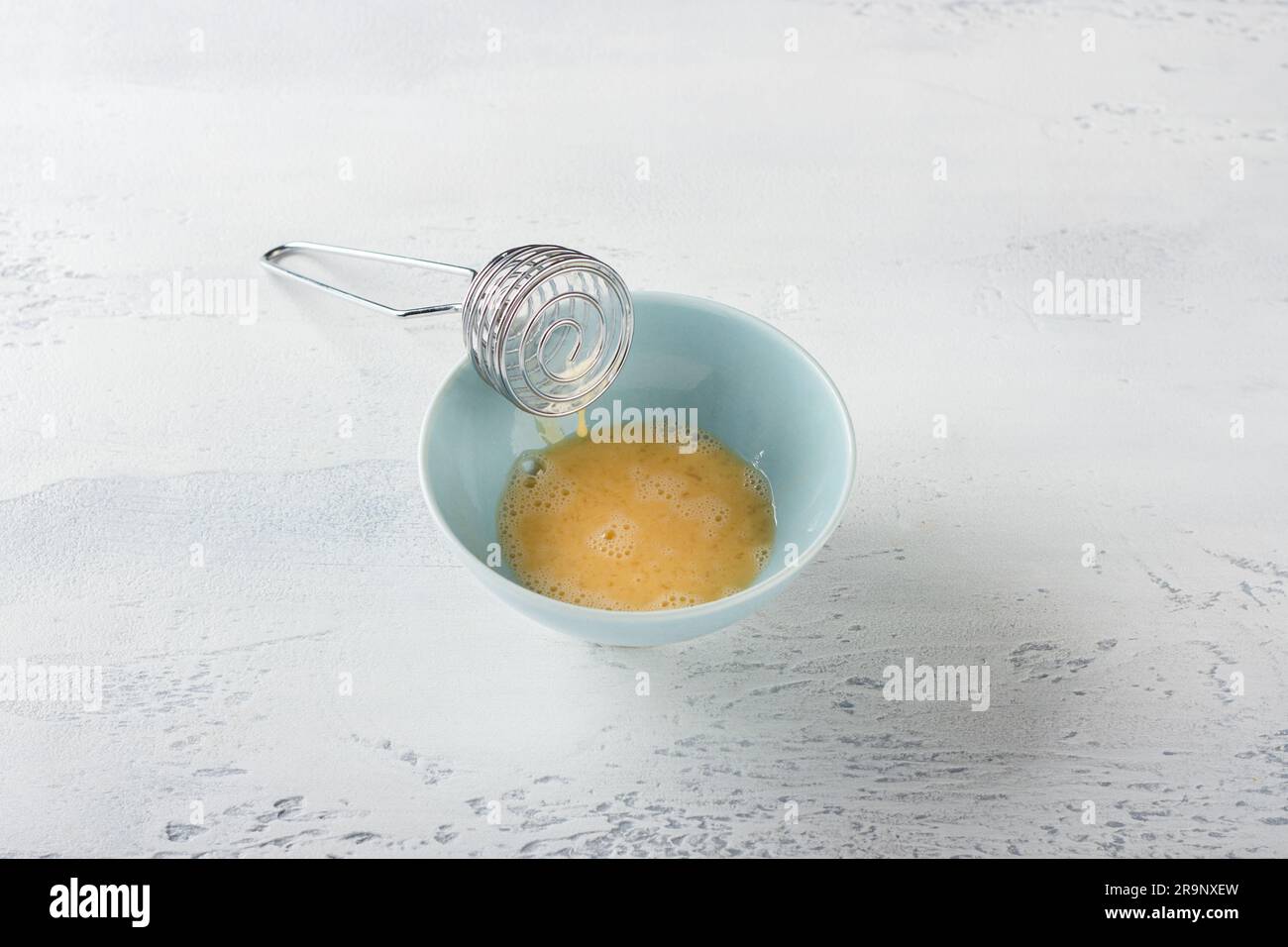 A blue bowl with a beaten egg and a whisk on a light blue table. Stage ...