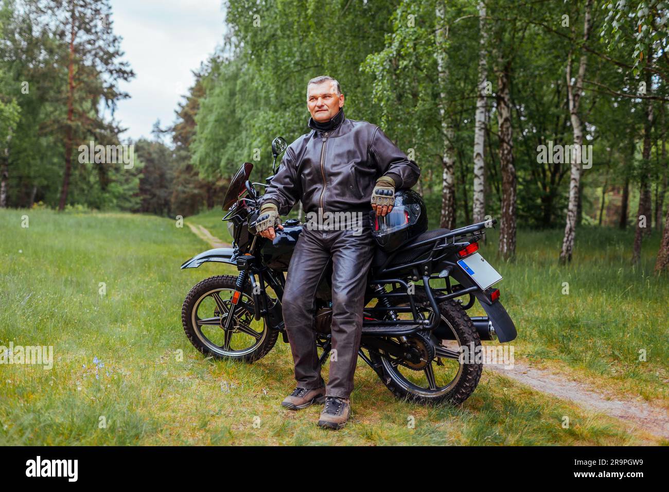 Elderly biker leaning on motobike in summer forest. Man in leather ...