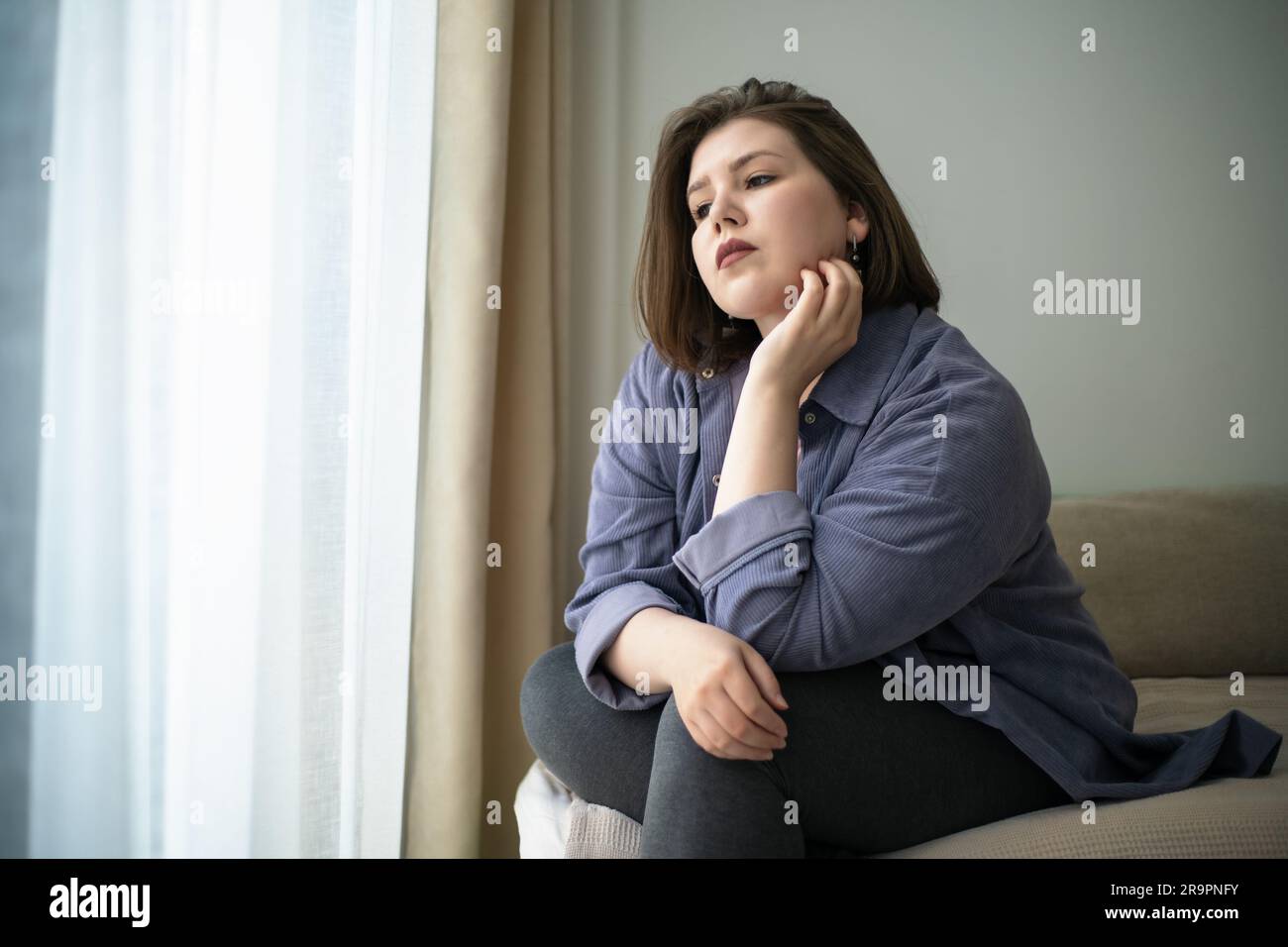Sad plump or overweight young girl looks out window portrait. Problems ...