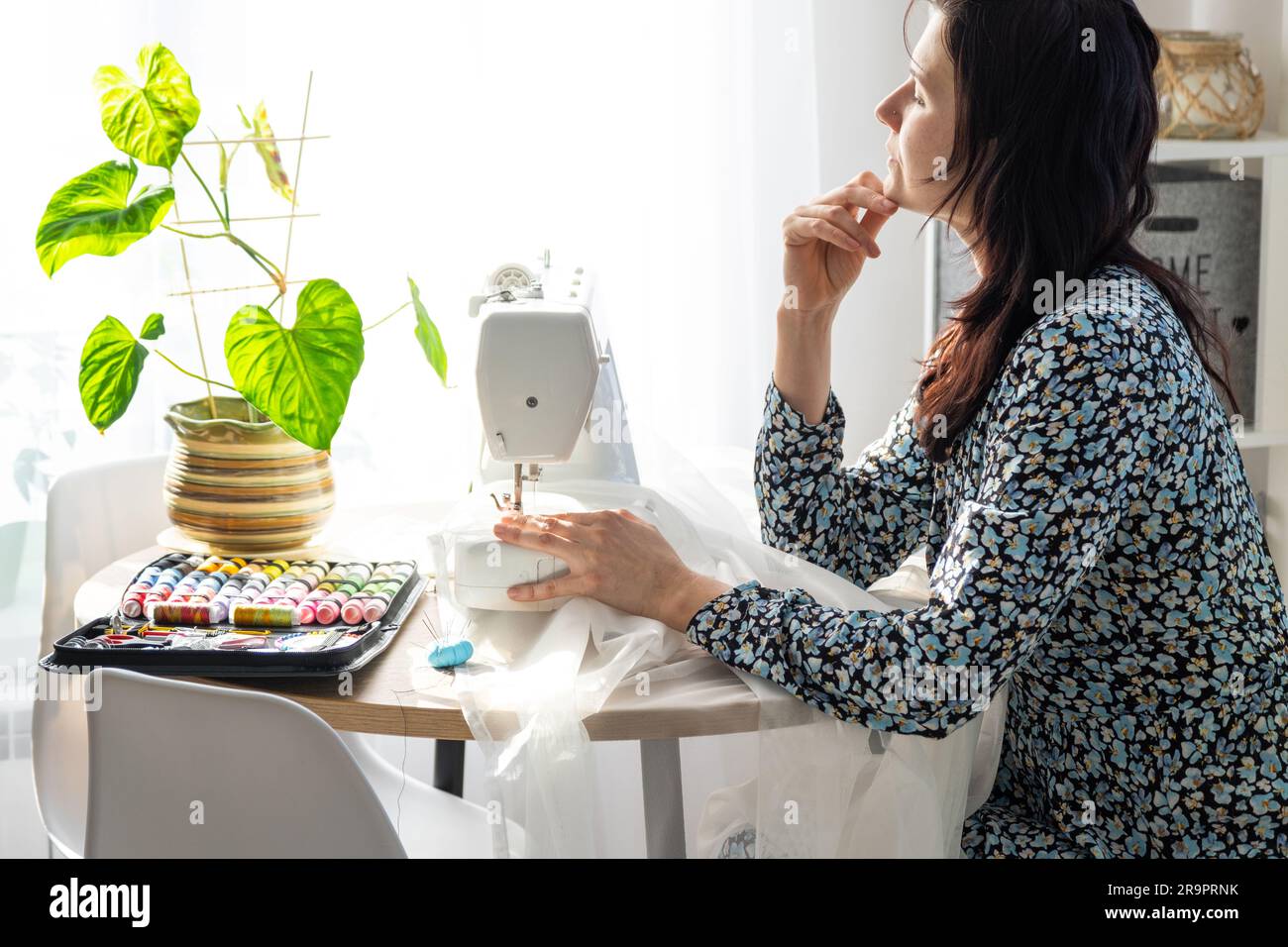Woman thinking, dreaming and sews tulle on an electric sewing machine in a white modern interior ...