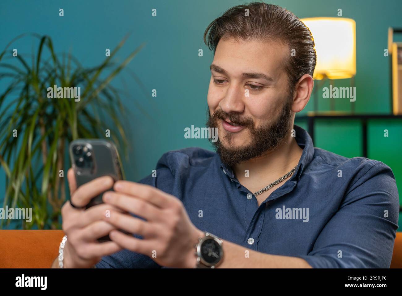 Lebanese man sits on sofa uses mobile phone smiles at home living room ...