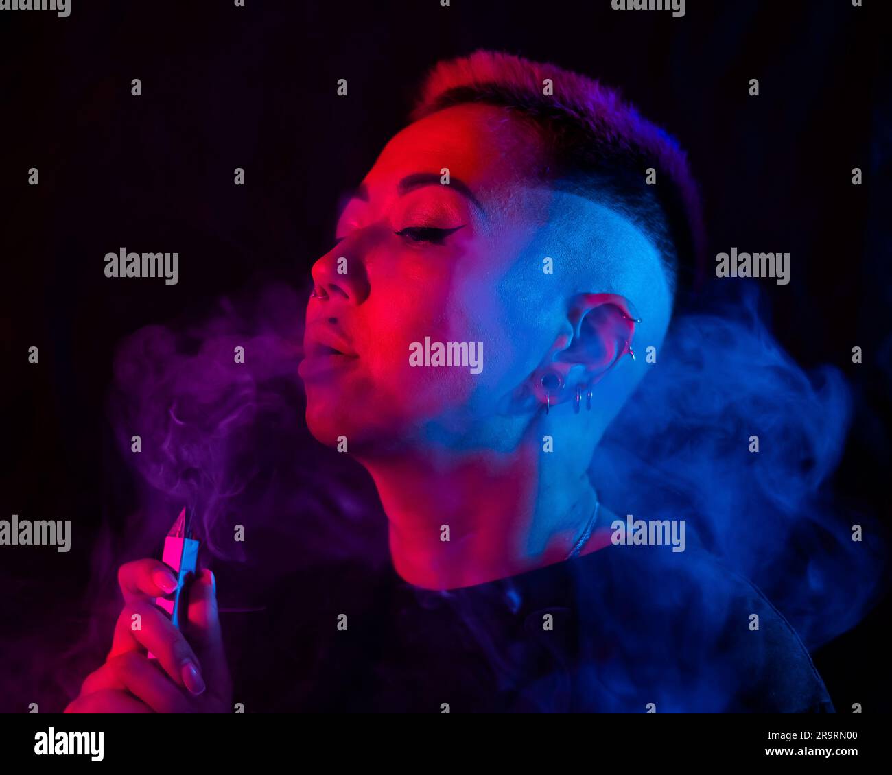 Portrait asian woman with shaved temples smokes vape in neon light ...