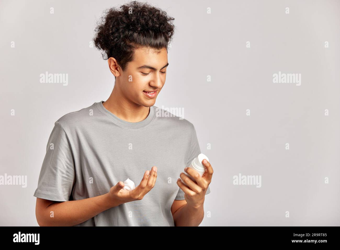 Young handsome smiling guy with black curly hair applying facial mask ...