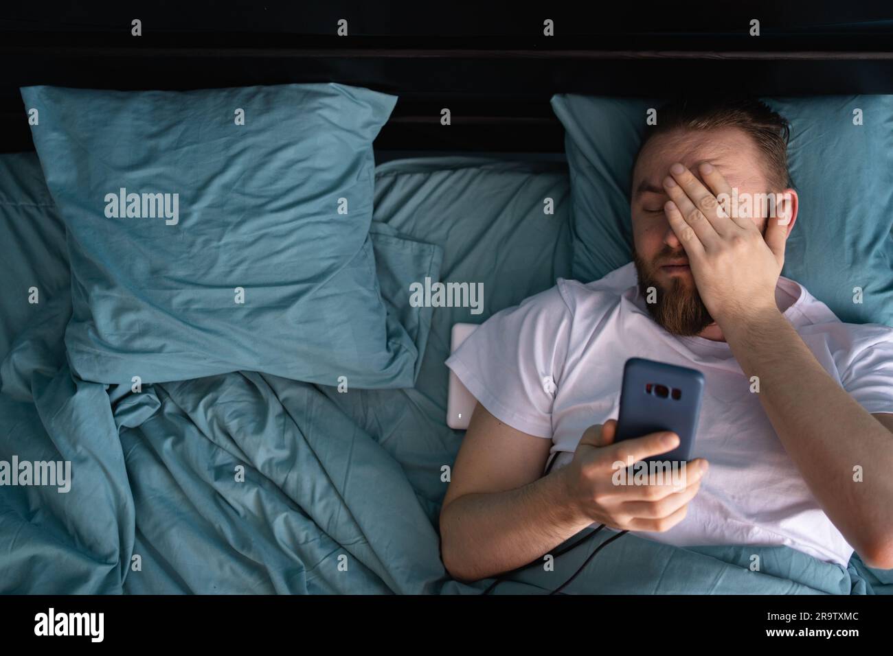 Confused man looking on mobile phone lying in bed at home in the ...
