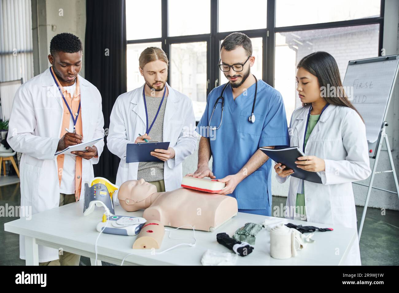 multiethnic students writing next to instructor showing wound care simulator near CPR manikin ...