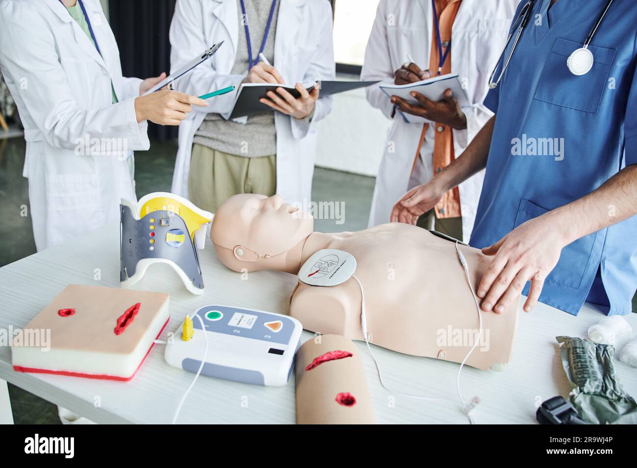 cropped view of woman in white coat pointing at CPR manikin and defibrillator near paramedic and ...