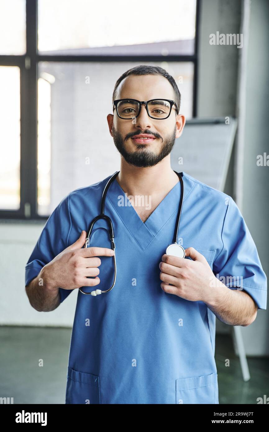 cheerful bearded healthcare worker with radiant smile standing in blue ...
