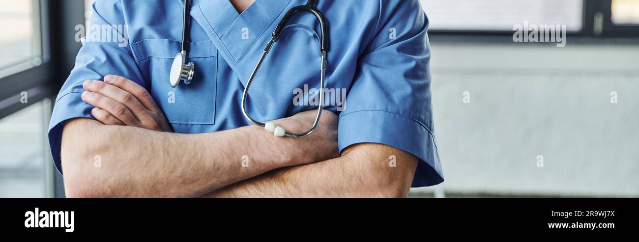 partial view of young doctor in blue uniform standing with crossed arms ...