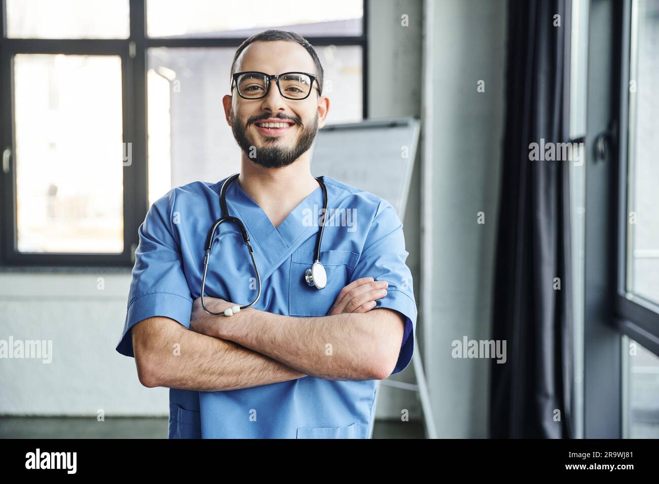 cheerful and bearded doctor in eyeglasses and blue uniform standing ...