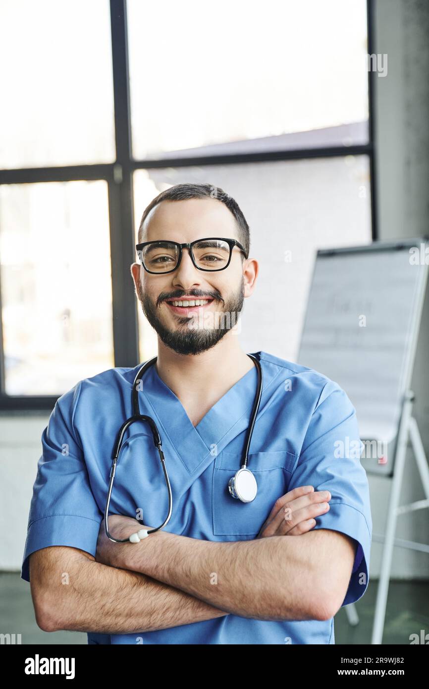 optimistic bearded paramedic in blue uniform and eyeglasses looking at camera and standing with ...