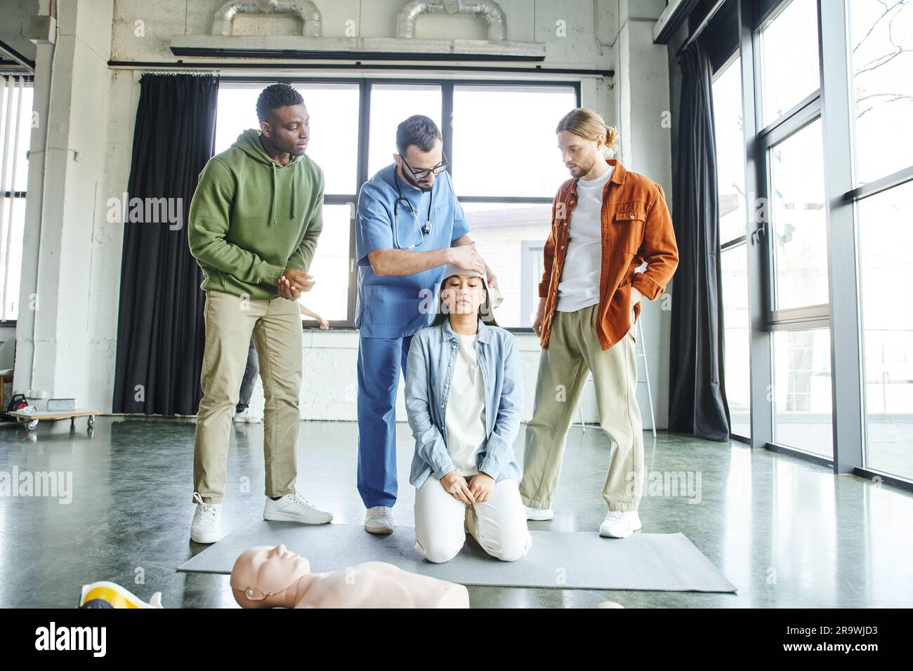 multiethnic man looking at professional paramedic bandaging head of asian woman near CPR manikin ...