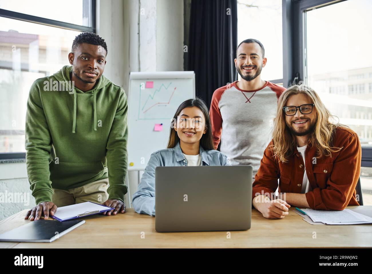 stylish and ambitious multicultural colleagues smiling at camera near laptop and flip chart with ...