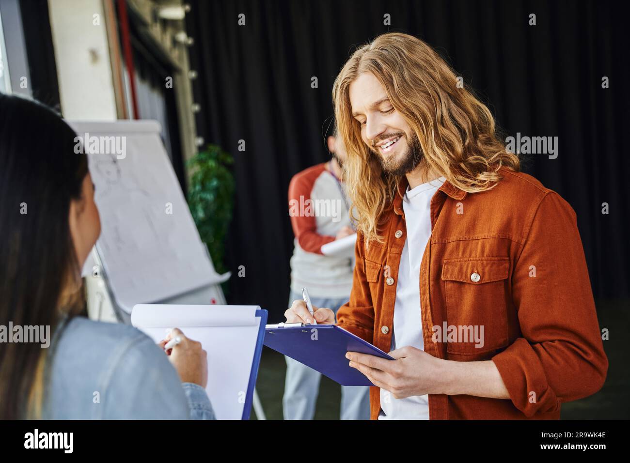 stylish and long haired man smiling while writing on clipboard near ...
